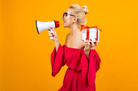 빨간 드레스를 입은 금발 소녀가 메가폰을 들고 있는 선물과 노란 배경에 손에 선물 상자를 들고 있는 것에 대해 이야기합니다 18 19세에 대한 스톡 사진 및 기타 이미지
