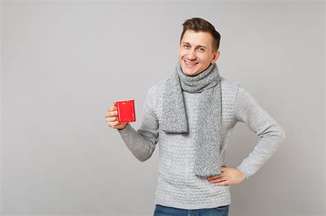 쾌활 한 젊은 남자가 회색 스웨터 스카프 커피 또는 차 스튜디오에 회색 배경에 고립의 빨간 컵을 들고 건강 한 생활 습관 병 아픈 질병 치료 추운 계절 개념 복사 공간을