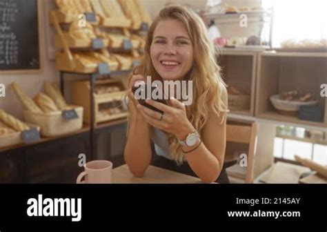Young Woman With Blonde Hair Smiling While Using A Smartphone In A Cozy Bakery Interior With