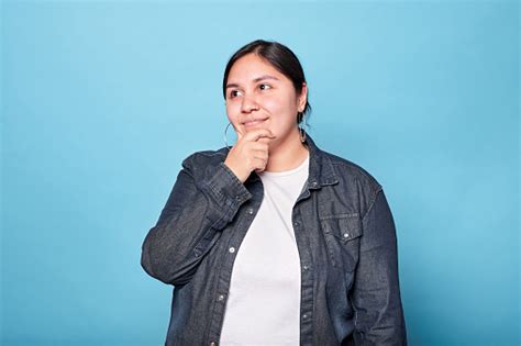 젊은 매력적인 라티 나 여자는 빈 공간을 찾고 턱에 손을 대고 데님 셔츠와 후프 귀걸이와 묶인 머리를 착용합니다 파란색 배경에