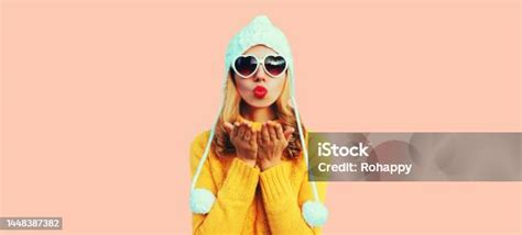 하트 모양의 선글라스 노란색 니트 스웨터 분홍색 배경에 폼폼이 있는 흰색 모자를 쓰고 달콤한 공기 키스를 보내는 그녀의 붉은 입술을 부는 아름다운 여성의 겨울 초상화 25