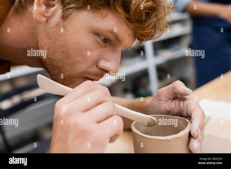 Concentrated Man With Red Hair Holding Shaper While Modeling Clay Cup In Pottery Workshop Stock