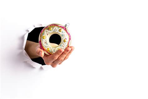 여성의 손은 장식 크리스마스 장난감을 뻗어 흰색 종이에 둥근 찢어진 구멍을 통해 화려한 유리 도넛 개념에 대한 스톡 사진 및