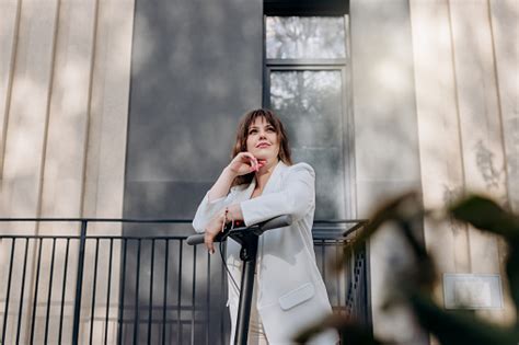 하얀 양복을 입은 아름다운 젊은 여성이 도시의 현대 건물 근처에서 전기 스쿠터와 함께 서서 멀리 바라 봅니다 30 34세에 대한