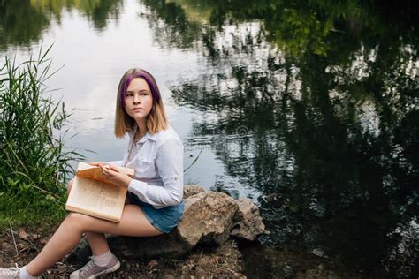 A Young Schoolgirl Sits On A Stone Near A Picturesque River With A Big Book In Her Hands Stock
