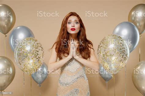 풍선 근처의 이브닝 드레스를 입은 젊은 빨간 머리 여성은 생일 파티 컨셉을 축하하는 평범한 파스텔 베이지 색 배경 스튜디오