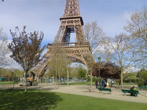 The Famous Eiffel Tower Among The Blue Sky Trees And Plants People And Park Benches To Sit In