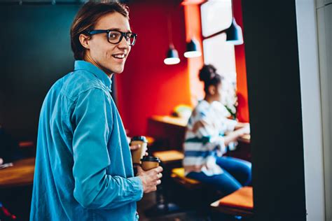 아침에 카페 인테리어에 시간을 보내는 여성 친구에게 커피를 들고 안경을 들고 있는 긍정적 인 백인 남자 카페 인테리어에서 음료와 함께 골판지 컵을 들고 있는 쾌활한 힙스터