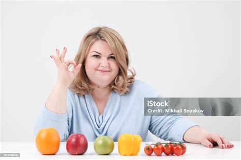 테이블에 흰색 바탕에 캐주얼 블루 옷에 젊은 매력적인 뚱뚱한 여자는 확인 야채와 과일은 행에 그녀의 앞에 배치 됩니다 보여줍니다 다이어트와 적절 한 영양 가냘픈에 대한 스톡