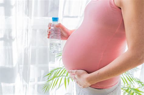 젊은 임산부는 운동 후 수분을 공급하기 위해 손에 물 한 병을 들고 창문 옆에 서 있습니다 임신 중 건강 관리 및 수분 공급의