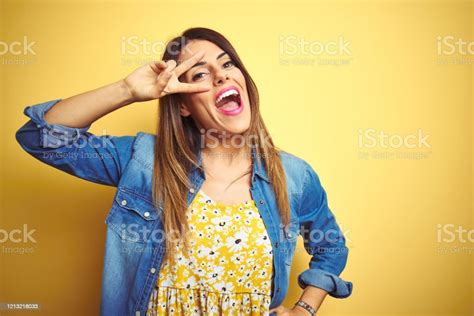 노란 고립 된 배경 위에 서있는 젊은 아름다운 여성은 얼굴에 손가락으로 평화 의 상징을하고 승리를 보여주는 쾌활한 미소 갈색
