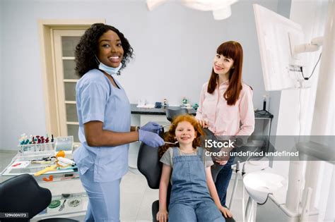 귀여운 십대 딸과 함께 웃고 있는 젊은 어머니가 소아 치과 의사인 아프리카 여성을 방문하여 Stomatology 클리닉에서 정기적인 치아 검진을 합니다 치과 의사의