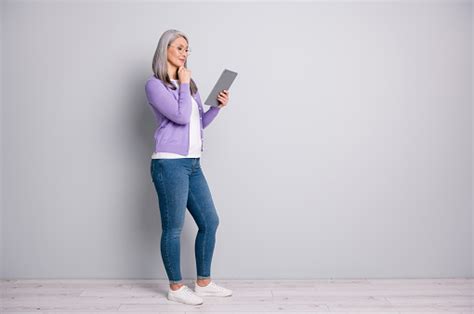 회색 색 배경에 고립 된 안경 캐주얼 의상을 입고 그녀의 얼굴을 만지는 노트북 독서와 함께 웃고있는 노인 여성의 전체 길이 신체 크기 사진 측면 보기에 대한 스톡 사진 및