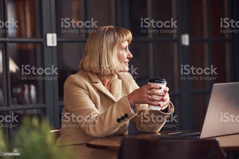 현대적인 노트북과 음료 한 잔을 손에 들고 카페실내에 앉아 있는 노인 여성의 초상화 60 64세에 대한 스톡 사진 및 기타