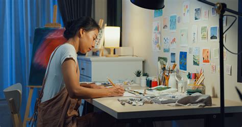 청소년 아시아 숙녀는 앞치마를 입고 책상 앞에 앉아 스케치북에 그림을 그리며 밤에는 집에서 아늑한 작업장으로 예술 작품을 만듭니다 현대 화가 추상 현대 미술 창의성 및