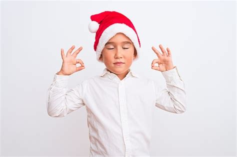 고립 된 흰색 배경 위에 서있는 크리스마스 산타 모자를 쓰고있는 아름다운 아이 소년은 손가락으로 명상 제스처를하고 눈을 감고 미소지으며 휴식을 취합니다 요가 개념입니다 12