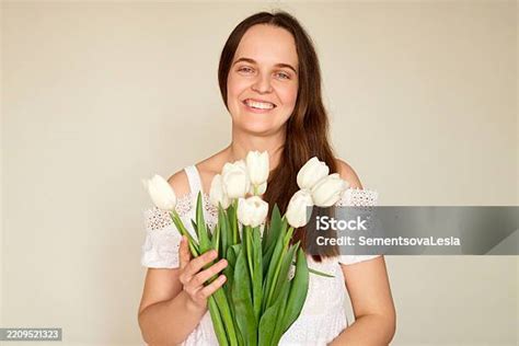 미소 짓는 여자가 새하얀 튤립 꽃다발을 들고 실내에 서 있습니다 하얀 드레스를 입고 긴 머리를 한 그녀는 기쁨과 아름다움의 순간을 포착합니다 30 34세에 대한 스톡 사진