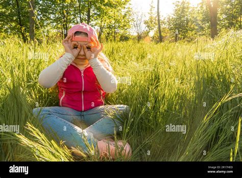 A Cheerful Girl Sits In The Thick Green Grass And Puts Her Palms To Her