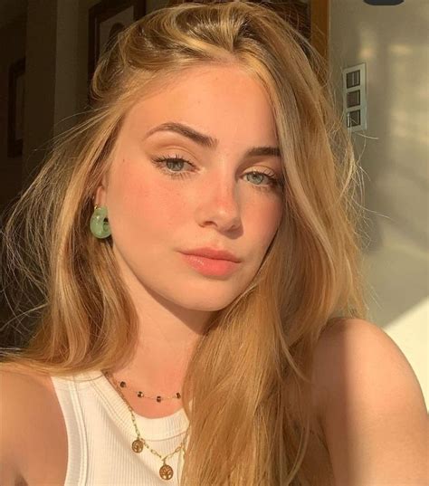 A Woman With Long Blonde Hair Wearing Green Earrings And A White Tank