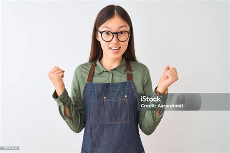 고립된 흰색 배경 위에 앞치마와 안경을 쓰고 있는 젊은 중국 상점 주인 여성은 팔을 들고 눈을 뜨고 성공을 축하하고 놀랐습니다