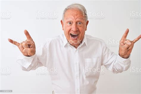 고립 된 흰색 배경 위에 서있는 우아한 셔츠를 입은 선임 회색 머리의 남자가 미친 표정으로 손을 들고 바위 기호를하고 있습니다
