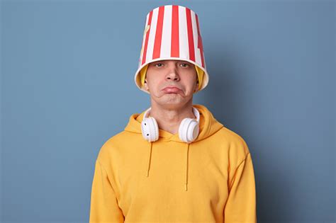 비니 모자와 캐주얼한 노란색 후드티를 입고 파란색 배경 위에 고립되어 서 있고 머리에 팝콘 양동이를 들고 포즈를 취하고 영화의