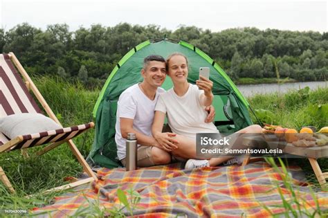 행복하고 즐거운 커플이 강 근처에서 시간을 보내는 사진 남자와 여자가 호수나 강가의 텐트 근처에 앉아 있고 스마트폰을 들고 셀카를 만드는 여성 2명에 대한 스톡 사진 및