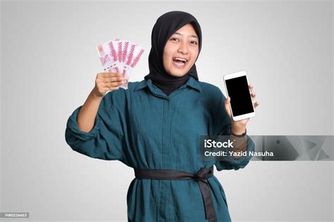 히잡을 쓴 흥분한 아시아 무슬림 여성의 초상화는 빈 화면 모형 휴대폰을 보여주면서 10만 루피아를 보여준다 재정 및 저축 개념 흰색 배경에 격리된 이미지 18 19세에 대한