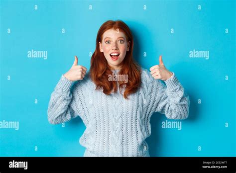 Amazed Redhead Girl In Sweater Showing Thumbs Up And Praising Product