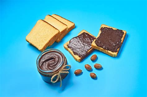 녹은 초콜릿이나 크림으로 가득 찬 유리 항아리는 바닐라 빵과 헤이즐넛 조각 근처에 부엌의 파란색 책상에 놓여 있습니다 영양가 있는 아침 식사 또는 점심 식사의 개념 가득