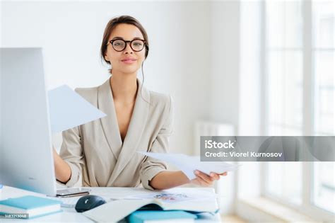 바쁜 성공한 여성 매니저는 사무실에서 서류를 가지고 일하고 데스크탑에서 포즈를 취하고 안경과 정장을 입고 보고서를 준비하느라