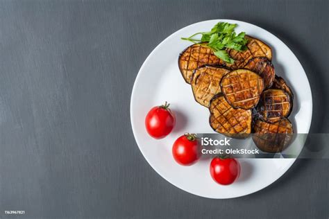 토마토와 파슬리를 전체 토마토와 파슬리로 장식 한 구운 가지 조각으로 가득 찬 흰색 세라믹 접시가 긁힌 어두운 콘크리트에 놓여
