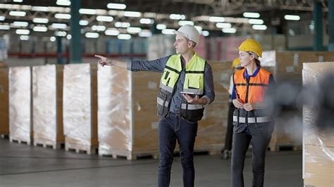 전문 남성 관리자 및 여성 부문 책임자는 하드 모자를 착용하고 제품 상자가있는 선반으로 가득 찬 소매 창고의 디지털 태블릿에 응용 프로그램을 통해 재고를 이야기하고 점검하며