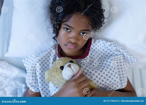 Portrait Of Sick Mixed Race Girl Lying In Hospital Bed Holding Teddy