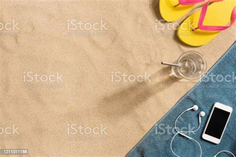 여름 방학 구성 플립 플롭 스마트 폰과 모래 배경에 물 한 잔 그림자가 있는 가혹한 빛 여름 배경입니다 수건으로 만든 국경