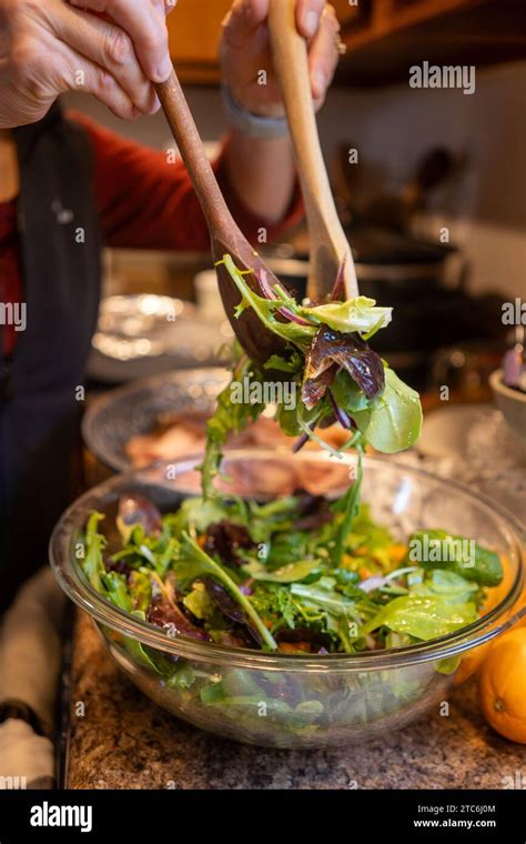 Asian Woman Tosses Lightly Dressed Salad With Wooden Spoons In Kitchen