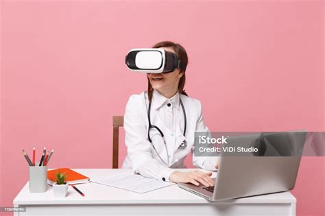 여성 의사는 파스텔 분홍색 벽 배경에 고립 된 병원에서 헤드셋 의료 문서와 컴퓨터에 책상 작업에 앉아 의료 가운 안경 청진에