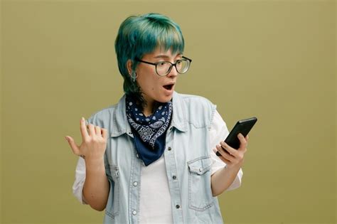 올리브 녹색 배경에 격리된 휴대전화를 사용하여 목에 안경 반다나를 착용한 놀란 젊은 여성 무료 사진