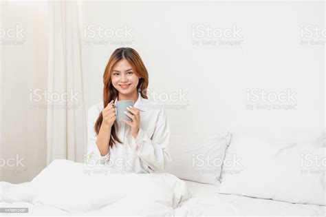 하얀 잠옷을 입은 여자가 주말 아침에 일어나 휴식을 취하고 노트북 휴대폰을 가지고 놀며 하얀 침실 안의 유리에서 빵을 먹고 차를