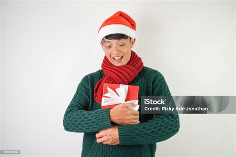 산타 클로스 모자 빨간 스카프 녹색 긴팔 스웨터를 입고 웃는 얼굴 표정을 가진 젊은 아시아 남자가 흰색 배경에 고립 된 리본이 달린 큰 선물 상자를 들고 있습니다 12월에