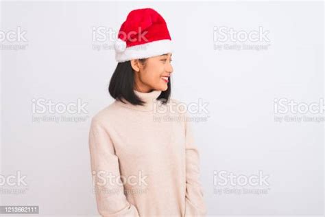 고립 된 흰색 배경 위에 크리스마스 산타 모자를 쓰고 있는 젊은 아름다운 중국 여성은 얼굴에 미소를 지으며 옆으로 바라보고