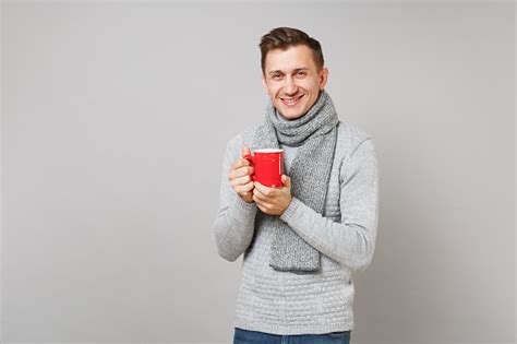 회색 스웨터에 젊은 남자가 미소 커피 또는 차 스튜디오에 회색 배경에 고립의 빨간 컵을 들고 스카프 건강 한 라이프 스타일 아픈 아픈 질병 치료 추운 계절 개념 복사 공간을