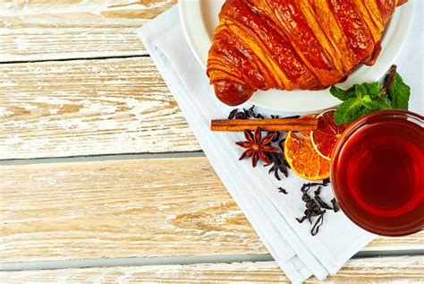 차가 있는 흰색 접시에 크루아상 오래된 나무 테이블에 차를 가진 머그잔 냅킨에 전체 홍차 레몬 민트 잎과 계피 음식과 가까운 텍스트를 위한 공간과 여유 공간을 복사합니다