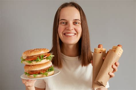흰색 티셔츠를 입은 검은 머리를 가진 웃는 쾌활한 젊은 성인 여성의 실내 사진은 큰 버거 샌드위치와 소시지를 반죽에 들고 패스트