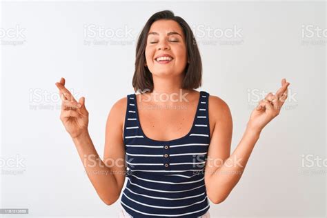 파란색 줄무늬 티셔츠를 입은 젊은 아름다운 여성은 고립 된 흰색 배경 몸짓 손가락 위에 서서 희망과 눈을 감고 미소를 지었습니다 행운과 미신 개념 T 셔츠에 대한 스톡 사진