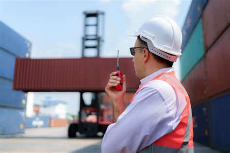 백인 의 선인은 컨테이너 창고 터미널에서 배경크레인 리프팅 컨테이너로 작업자를 명령하기 위해 무전기를 사용하고 있습니다 35 39세에 대한 스톡 사진 및 기타 이미지