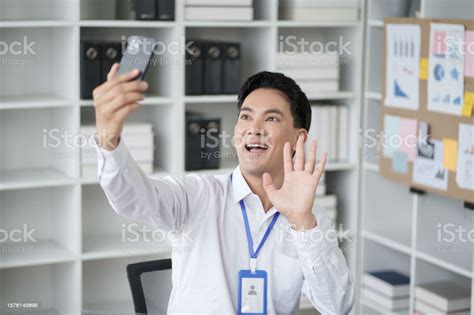 쾌활한 젊은 아시아 사업가가 사무실에 앉아 친구들과 화상 통화를 하고 있다 Brand Name Smart Phone에 대한 스톡