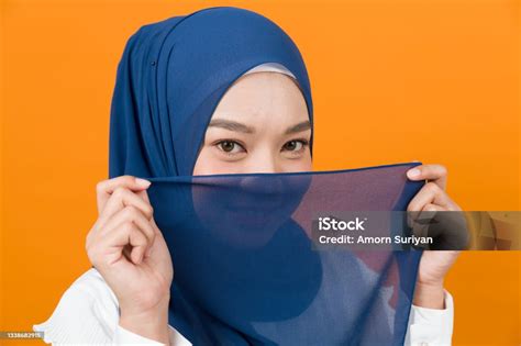 히잡 머리 스카프를 들고 있는 아시아 의 젊은 무슬림 여성이 그녀의 얼굴을 덮고 있다 스튜디오에서 오렌지 색 배경에 히잡 머리