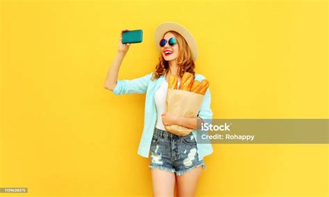 초상화 행복 한 웃는 여자 긴 흰색 빵 바게트 짚 모자 화려한 노란색 배경에 반바지를 착용 종이 가방을 들고 휴대 전화에 의해