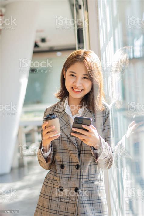 젊은 아시아 사업가들은 커피 한 잔과 스마트폰을 들고 사무실의 유리벽 옆에 서 있었다 Brand Name Smart Phone에